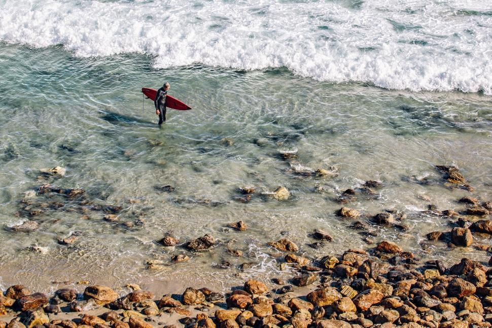 Free Stock Photo of A surfer with a red board stands on a rocky ...